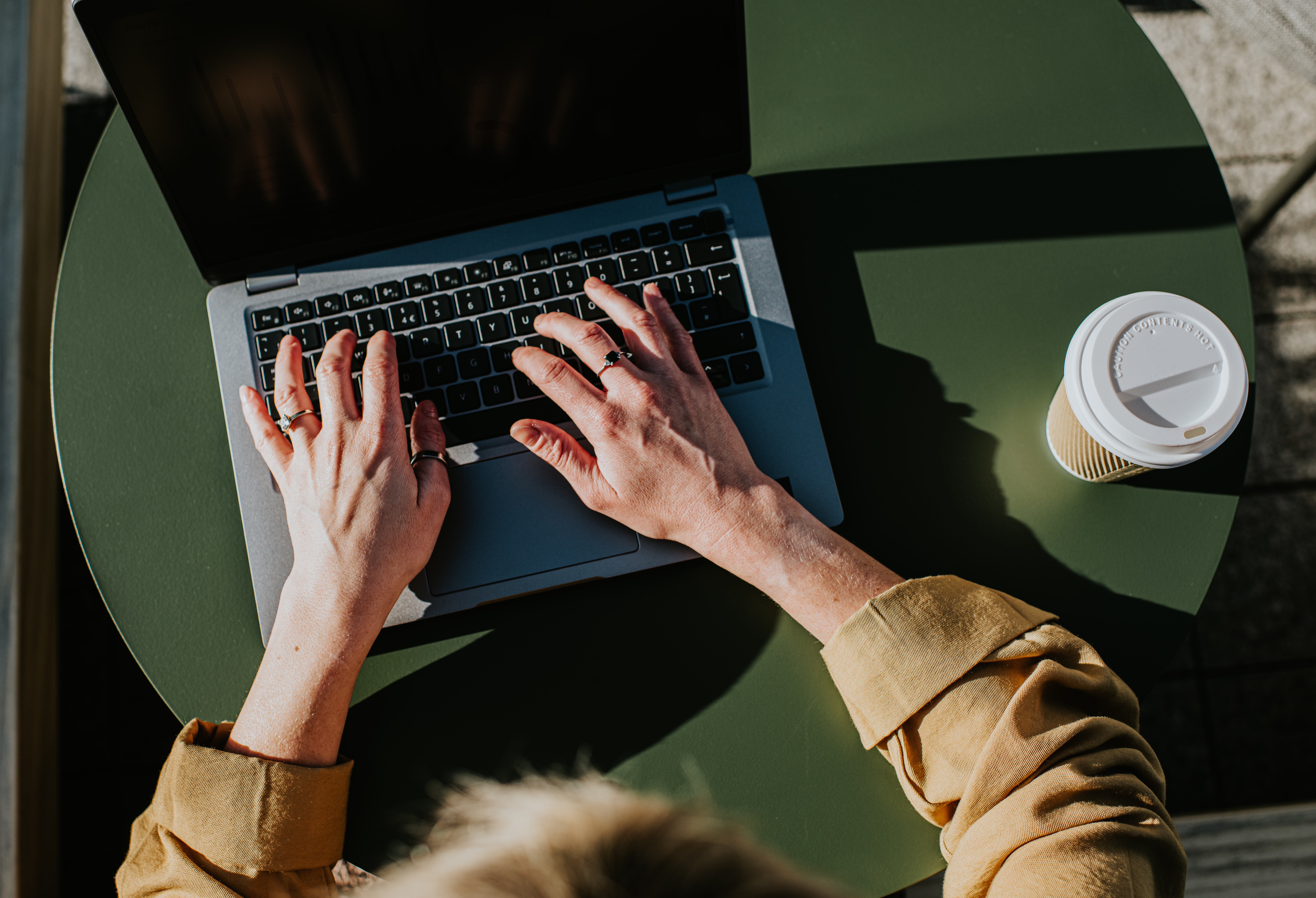 Hands typing on a laptop at an outdoor table with a coffee cup.